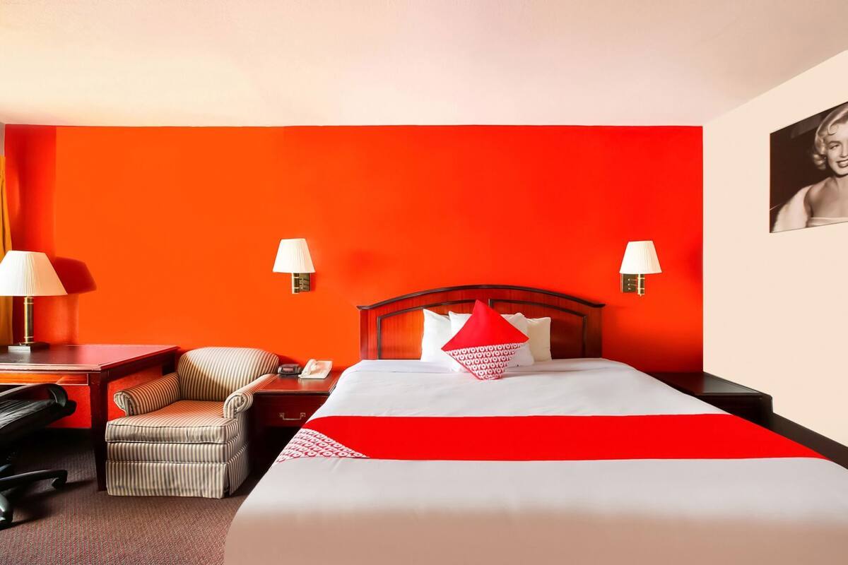 Quarto de hotel com uma cama, uma escrivaninha, uma cadeira e duas luminárias fixadas na parede.