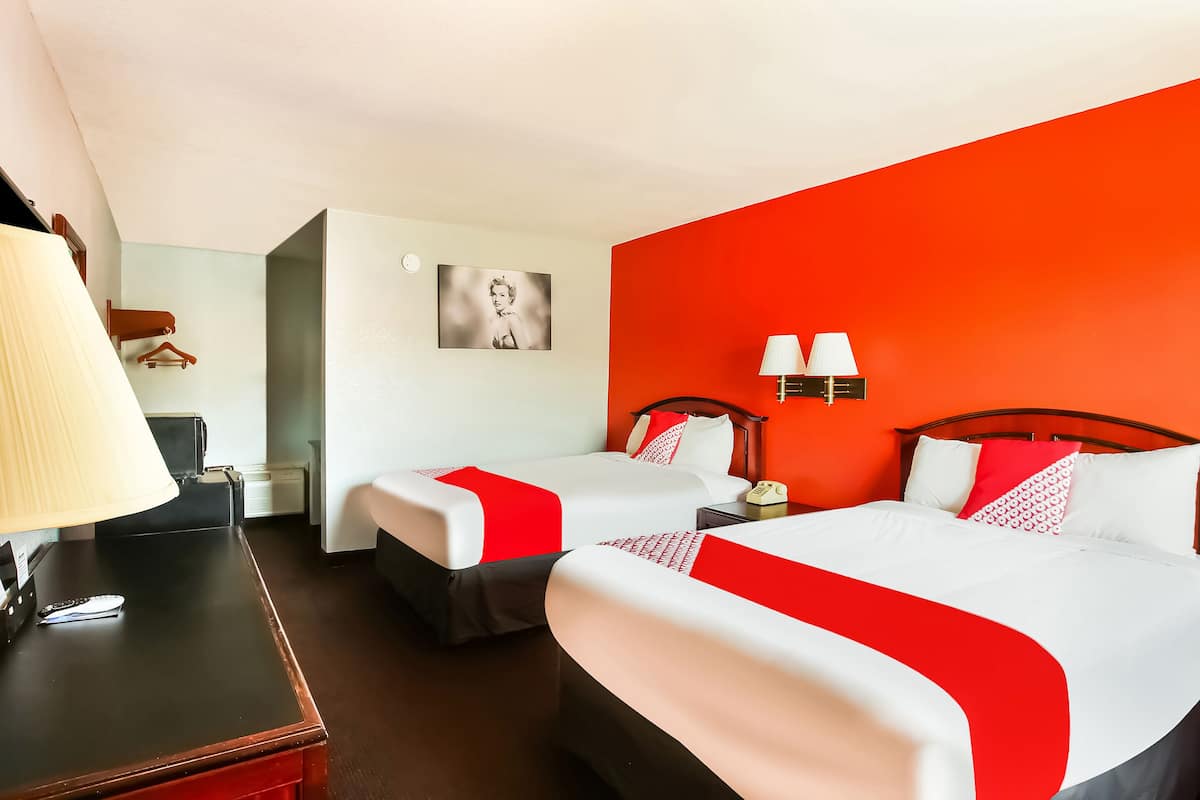 Quarto de hotel com duas camas, uma mesa de cabeceira, uma luminária e uma parede com detalhes em vermelho.