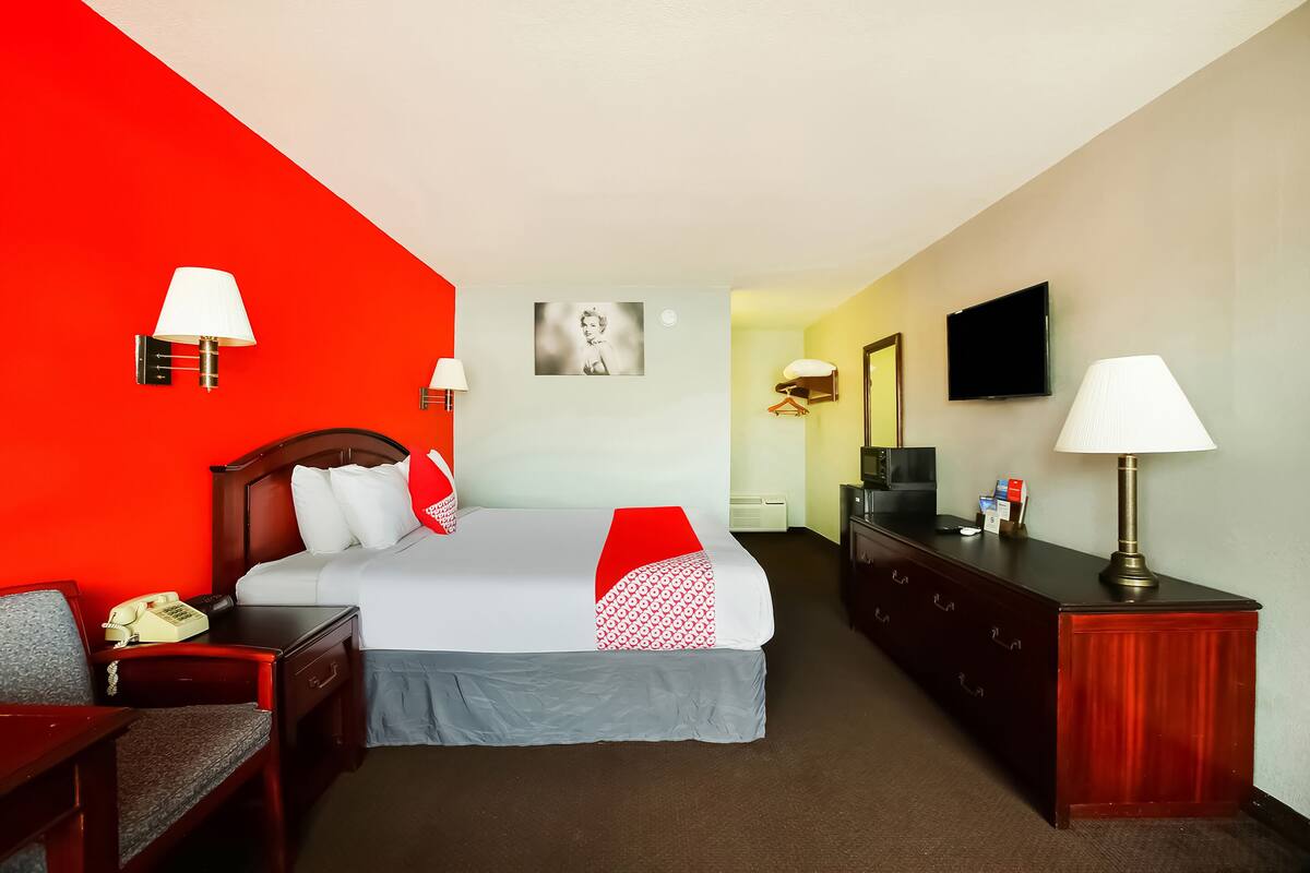 Quarto de hotel com cama, mesa de cabeceira, abajur, cômoda e televisão.