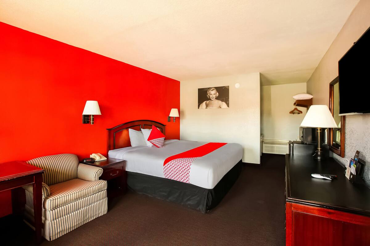 Quarto de hotel com uma parede em vermelho, cama com colcha branca e vermelha, poltrona listrada, escrivaninha com abajur e televisão.