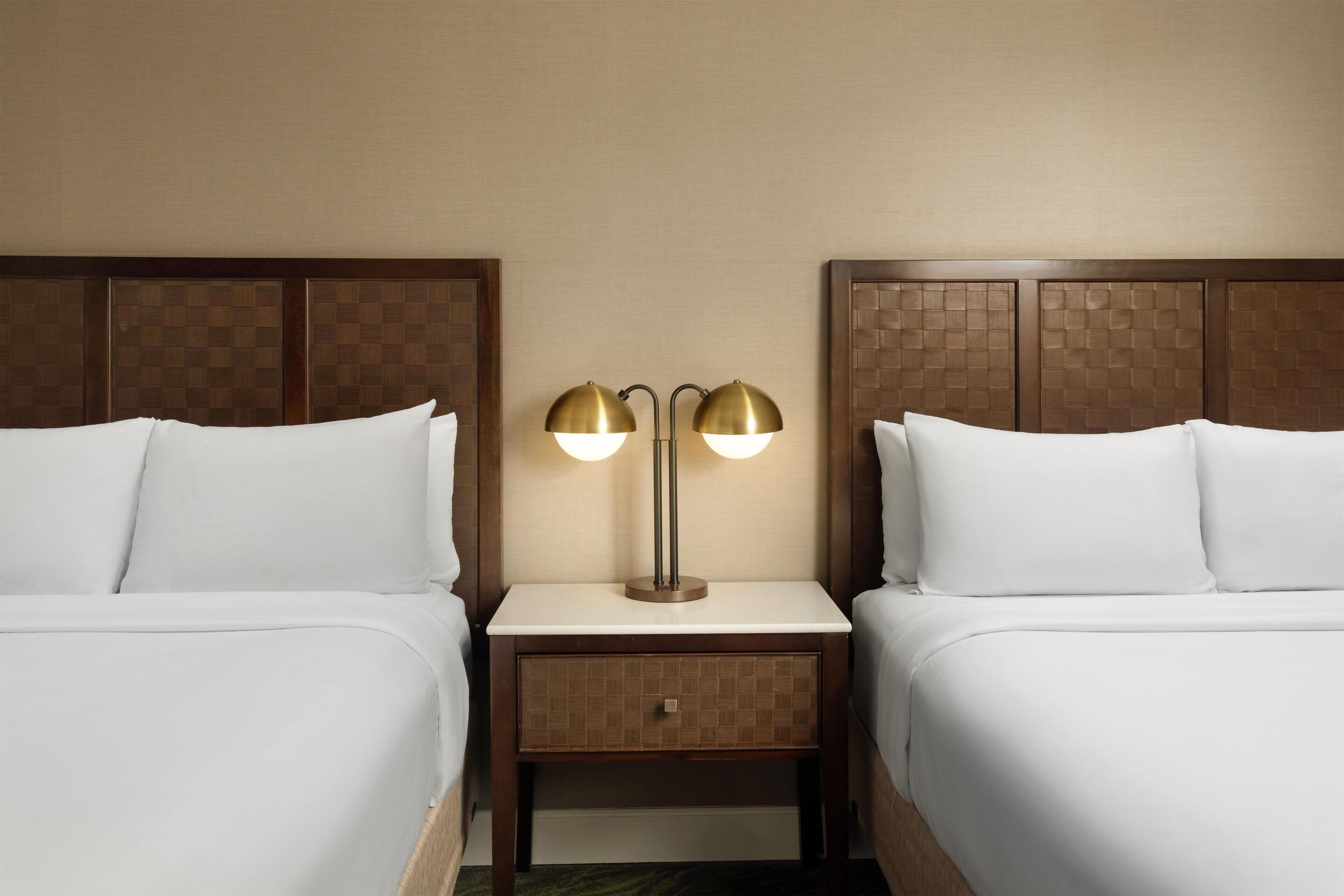Quarto de hotel com duas camas, uma mesa de cabeceira com abajur e cabeceira de madeira.
