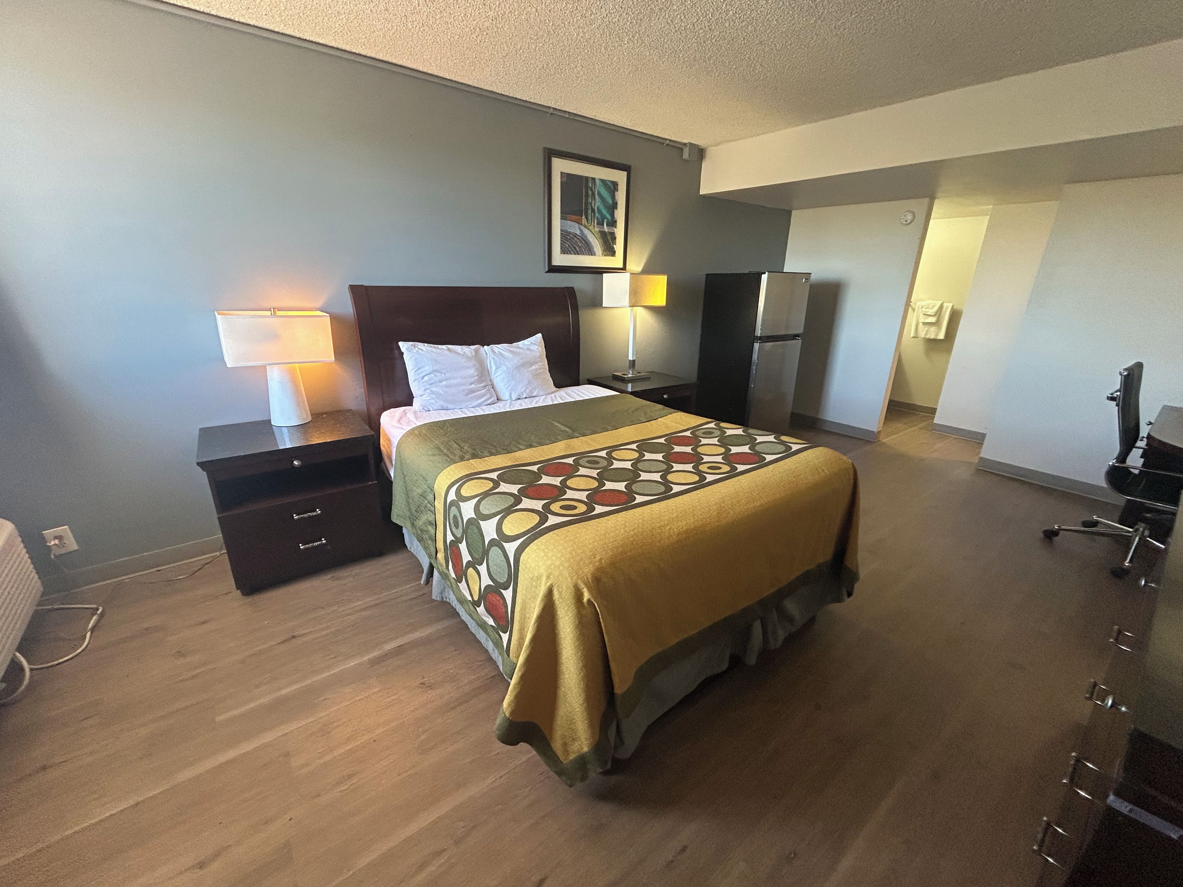 Quarto de hotel com cama, mesas de cabeceira, luminária, geladeira e escrivaninha com cadeira.