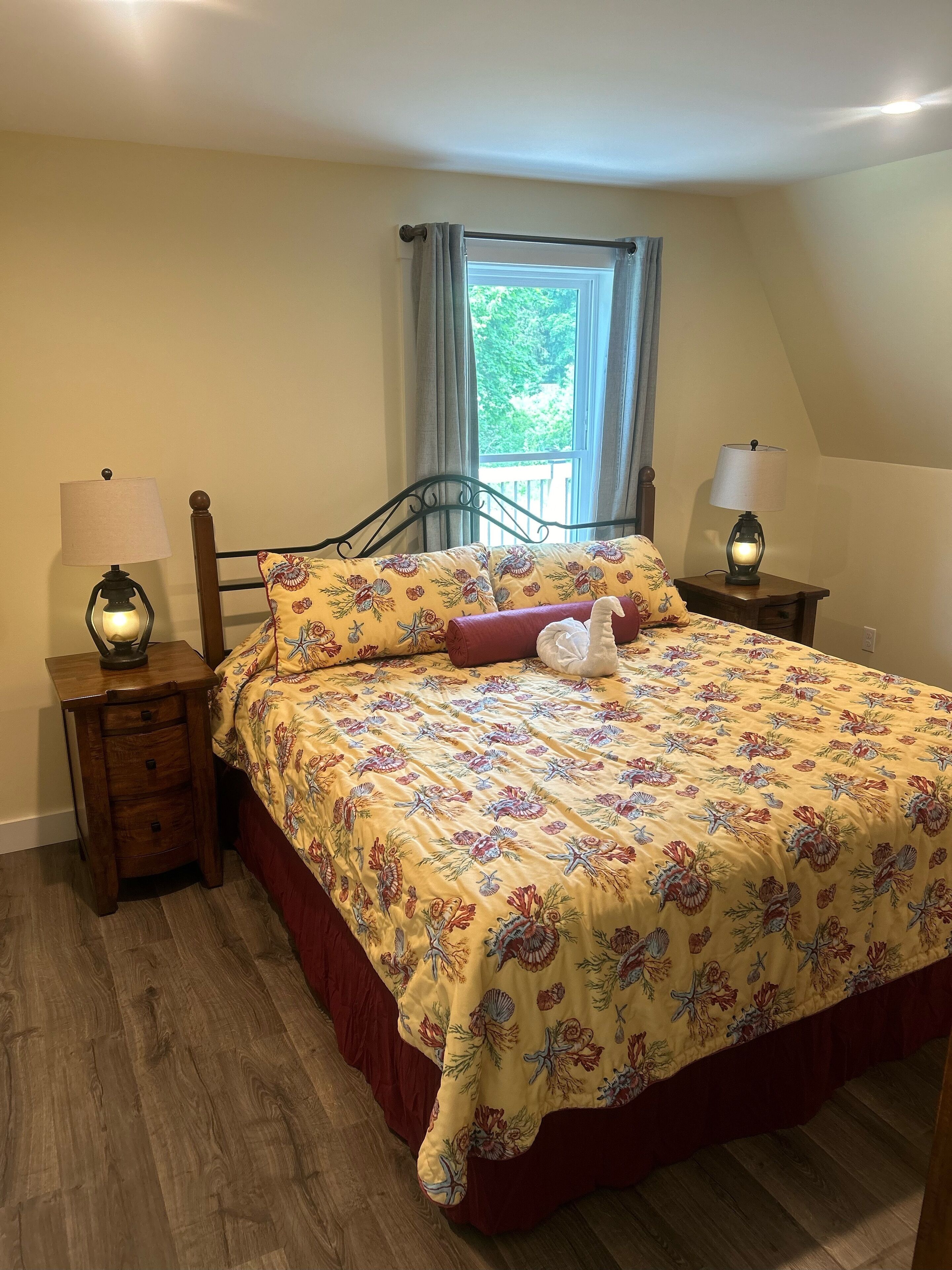 Quarto com cama, duas mesas de cabeceira, janela com cortinas e vista para o verde lá fora.
