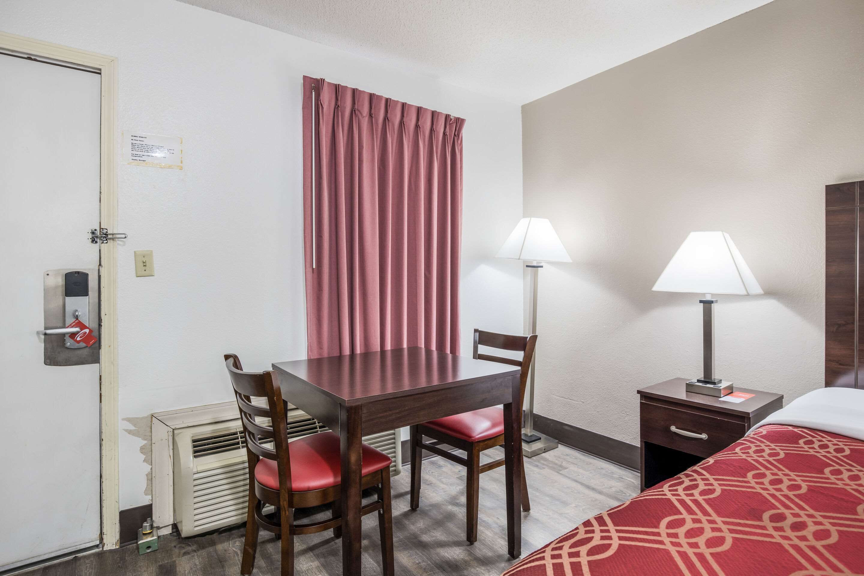 Quarto de hotel com mesa de madeira, duas cadeiras, cama com colcha estampada, mesa de cabeceira com abajur e janela com cortina vermelha.