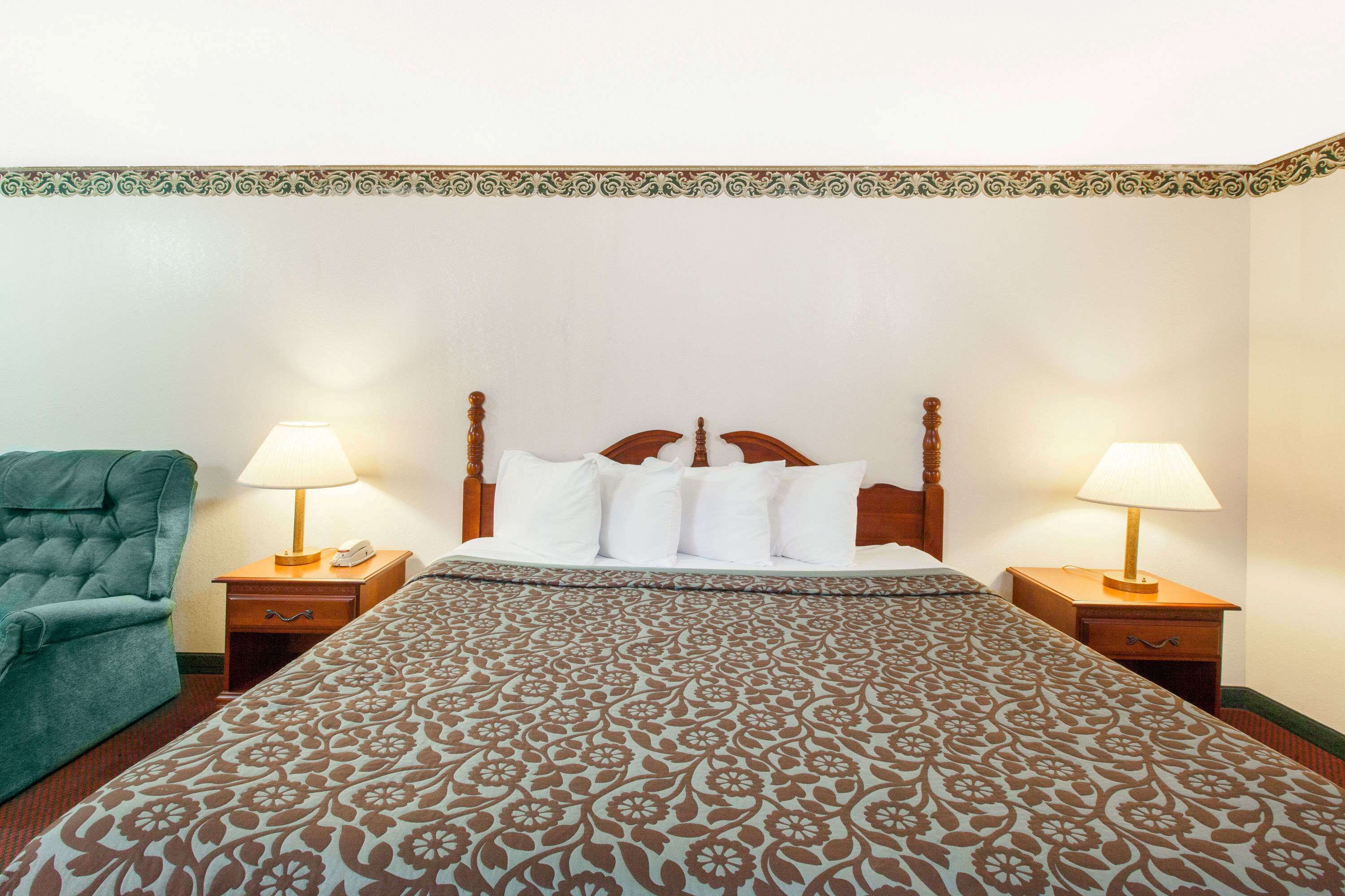 Quarto de hotel com cama, duas mesas de cabeceira com luminárias, uma cadeira e um detalhe decorativo na parede.