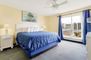 Quarto de hotel com uma cama grande, duas luminárias de cabeceira, um ventilador de teto, uma janela com cortinas e um quadro na parede.