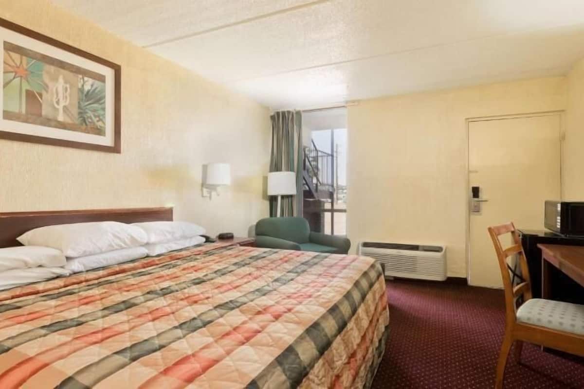 Quarto de hotel com cama, cadeira, escrivaninha com televisão, janela com cortinas e um quadro na parede.