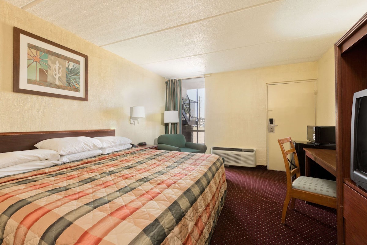 Quarto de hotel com uma cama grande, uma escrivaninha com televisão, uma cadeira, uma janela com cortinas e um quadro na parede.