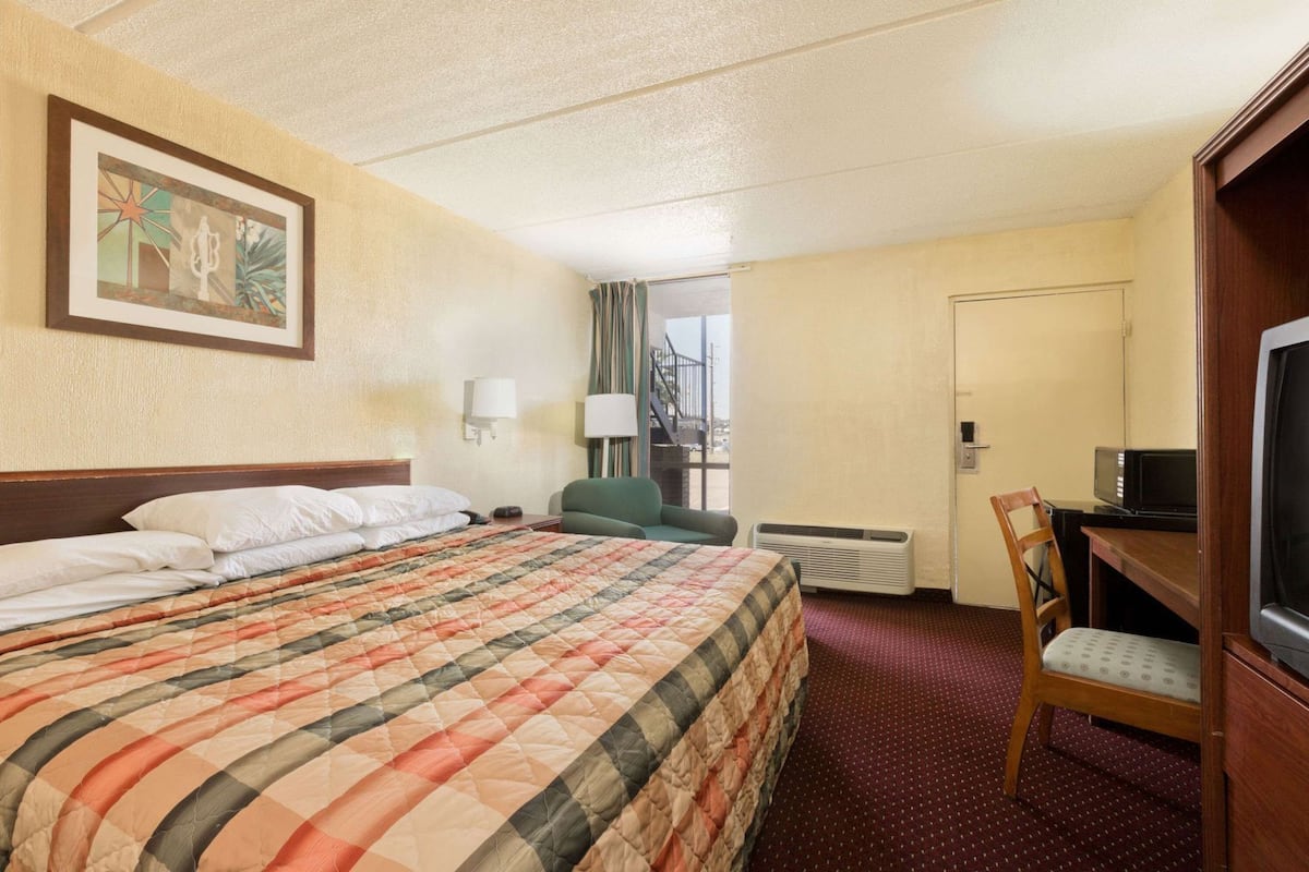 Quarto de hotel com cama, escrivaninha com televisão, cadeira, janela com cortinas e um quadro na parede.