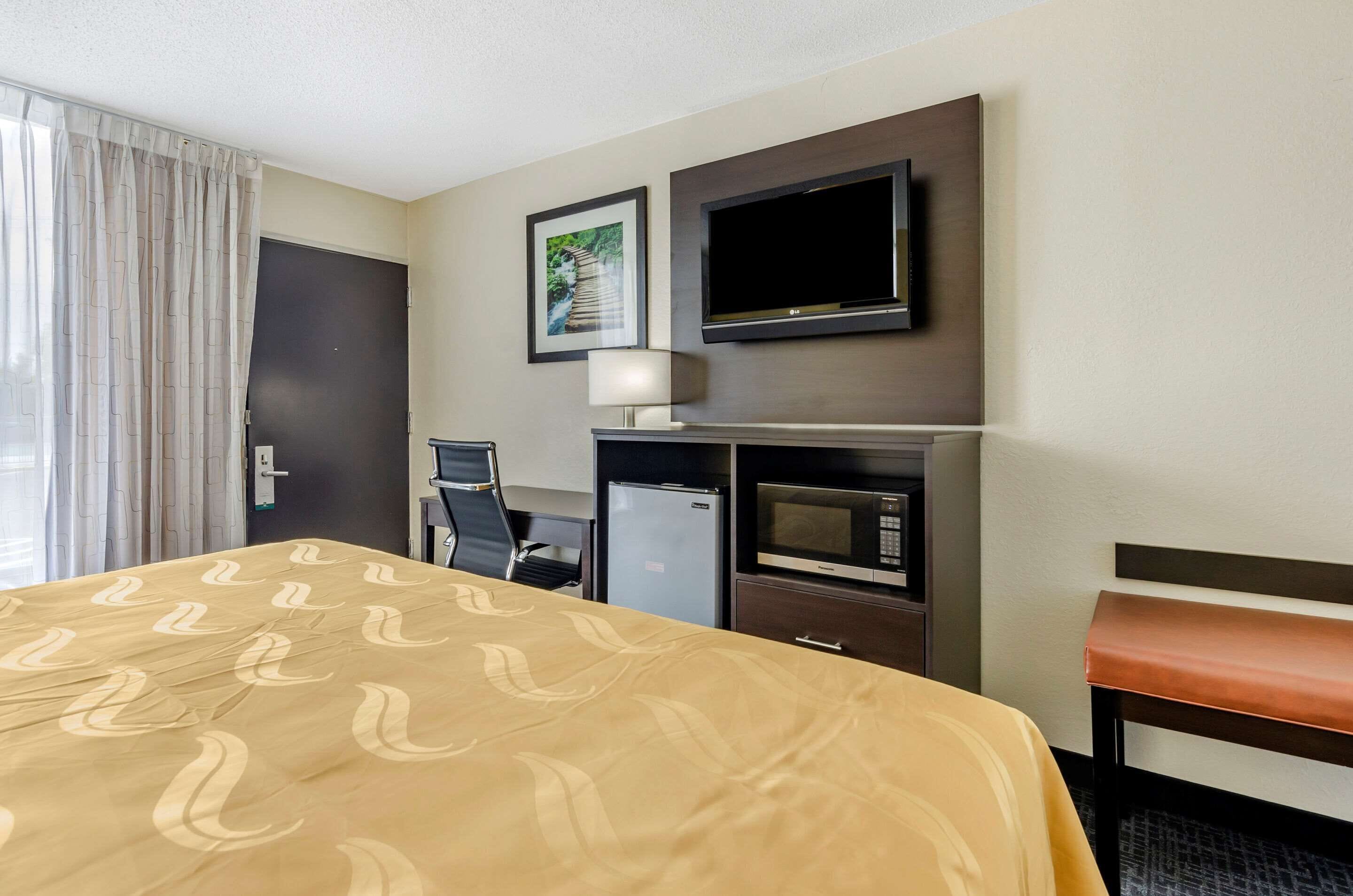 Quarto de hotel com cama, escrivaninha com cadeira, televisão, micro-ondas e frigorífico.