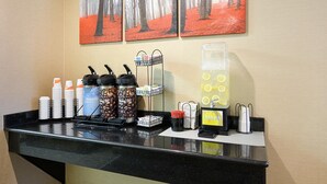 Uma área de recepção de hotel com balcão preto, dispensadores de café e suco, e uma pequena geladeira.