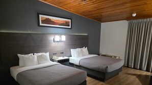 Quarto de hotel com duas camas, um quadro pendurado, uma mesa de cabeceira com telefone e uma janela com cortinas.