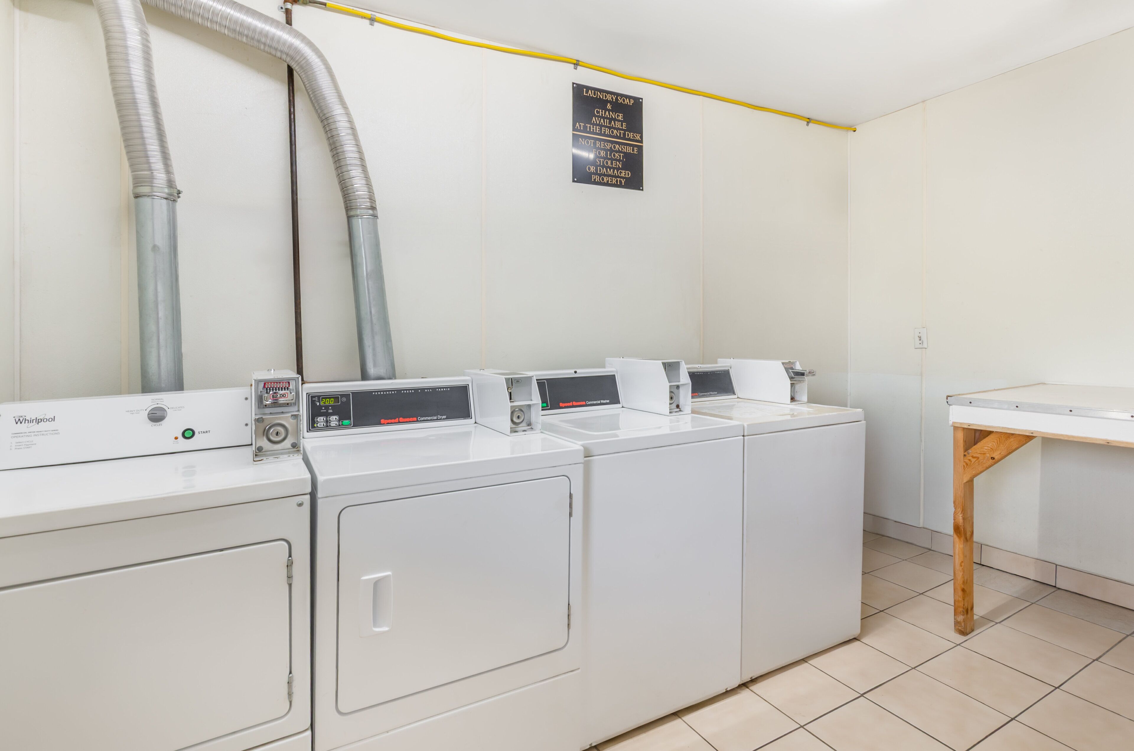Uma lavanderia com quatro máquinas de lavar e secadoras Whirlpool, um aviso sobre sabão para roupa e uma mesa de madeira.