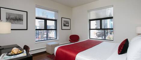 Quarto de hotel com uma cama grande, uma escrivaninha com cadeira, uma cadeira vermelha e duas janelas com vista para a cidade.