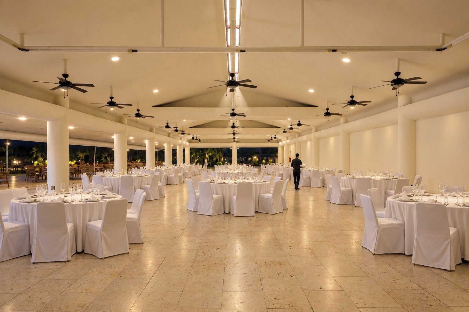 Amplo salão de banquetes coberto, com várias mesas redondas dispostas para refeições, ventiladores de teto e um layout espaçoso.