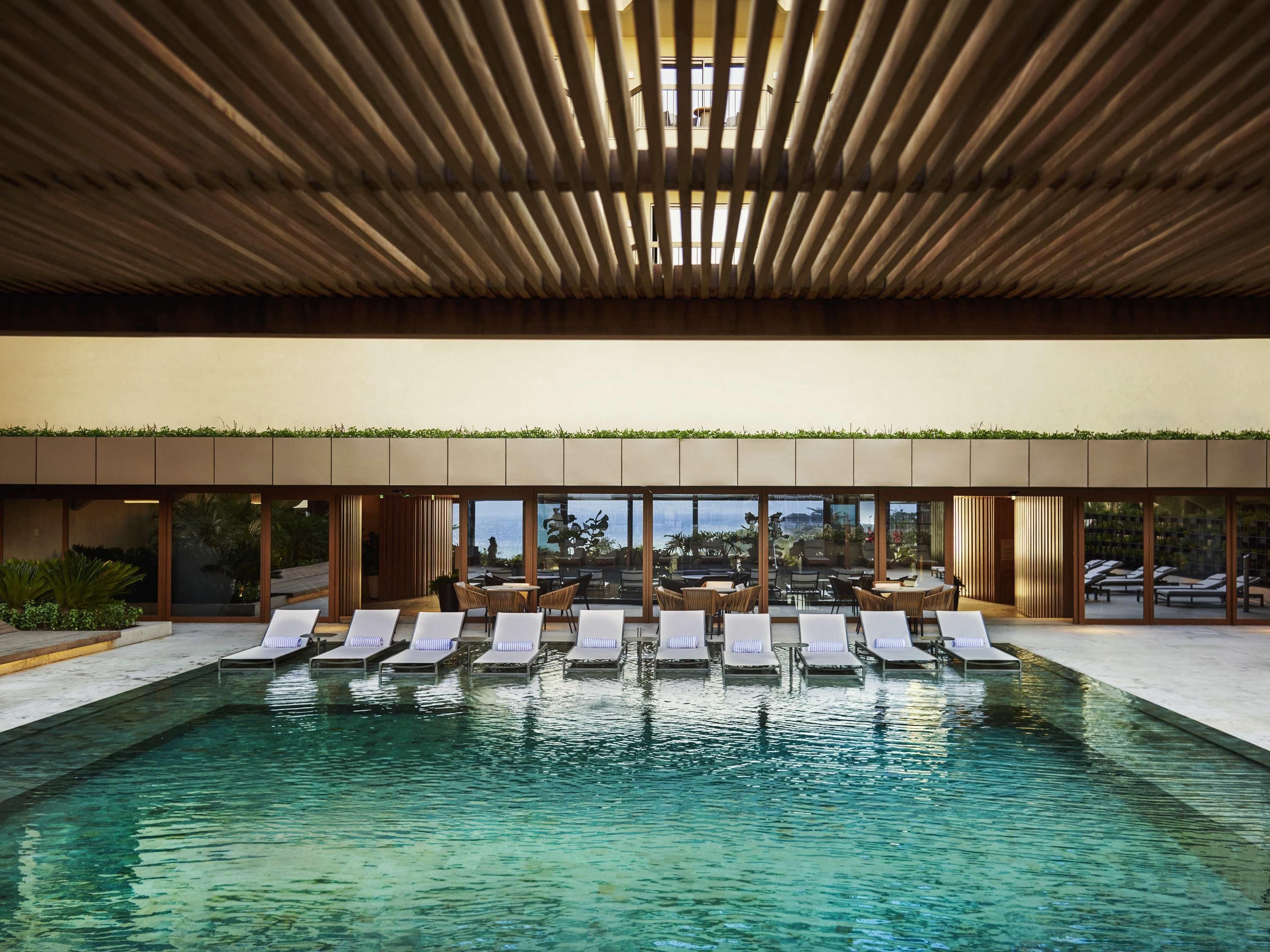Uma área interna de piscina moderna, com água cristalina, espreguiçadeiras e vista para o mar através de grandes janelas.
