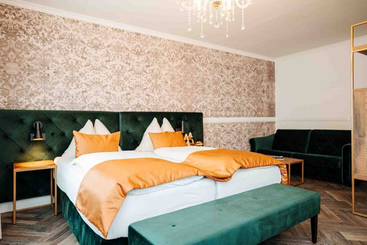 Quarto com cama grande, banco estofado verde, papel de parede estampado e lustre.