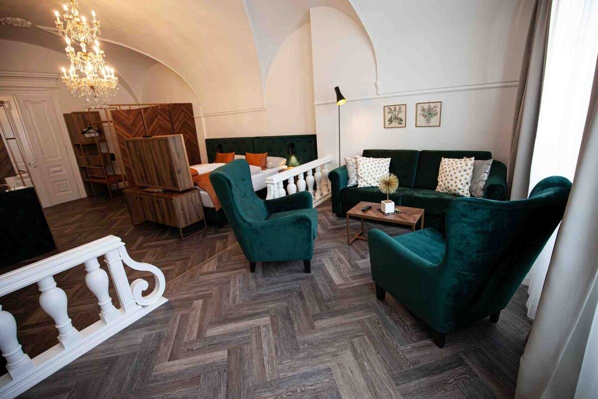 Uma sala de estar ampla com piso em padrão herringbone, um sofá verde, uma mesa de centro de madeira e um lustre.