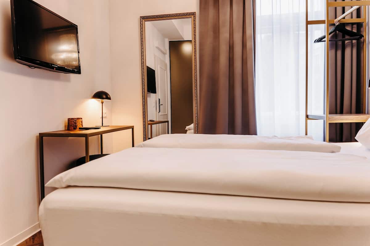 Quarto de hotel com uma cama, televisão presa à parede, mesa de cabeceira com abajur, espelho e guarda-roupa com roupas penduradas.