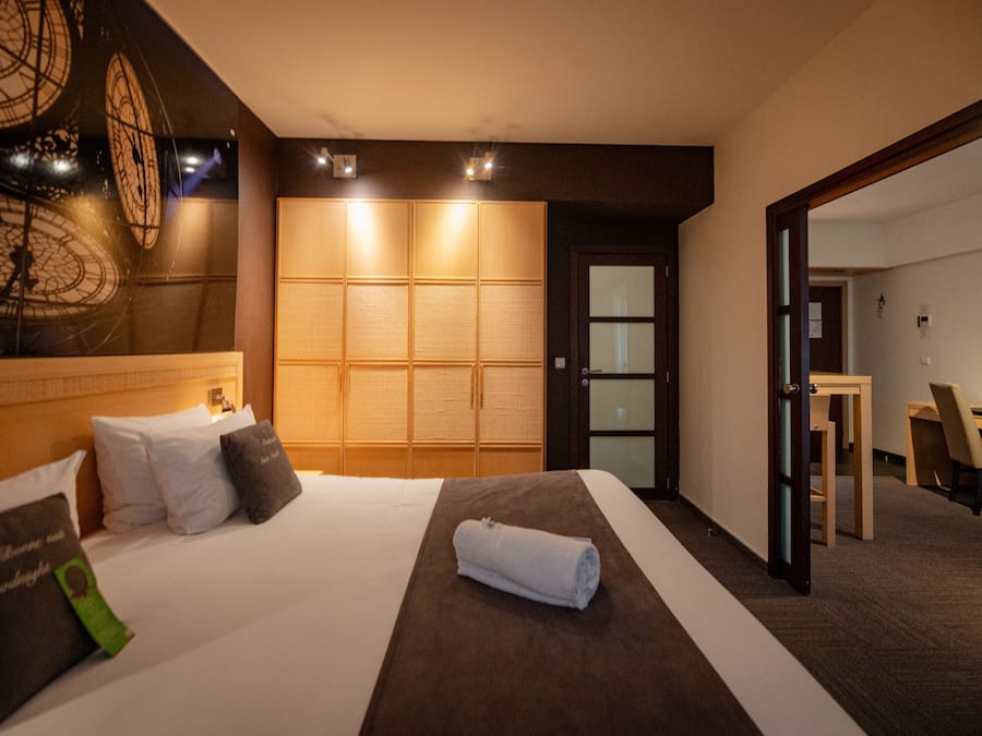 Quarto de hotel com uma cama grande, um guarda-roupa de madeira, uma escrivaninha com cadeira e um espelho.