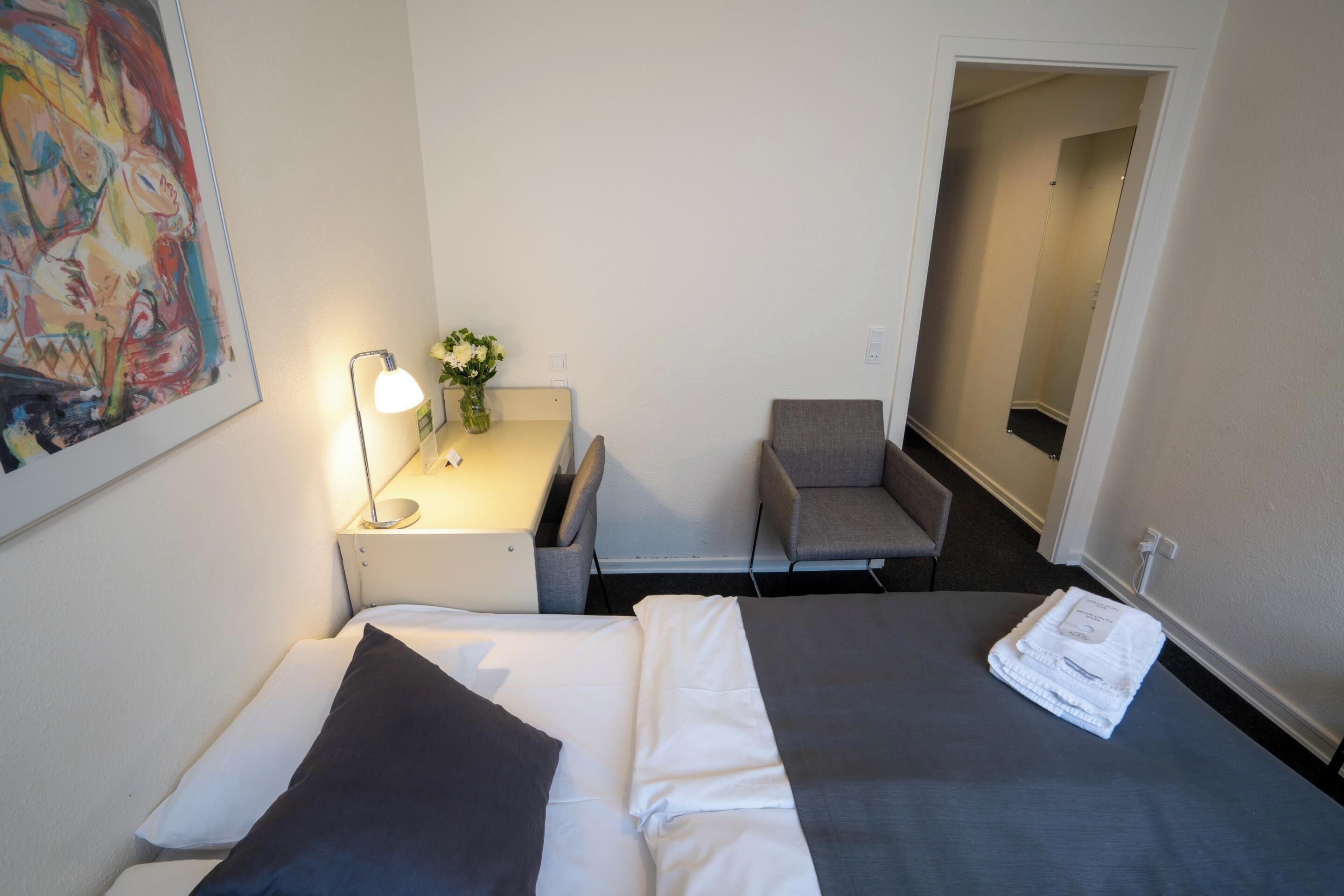 Quarto de hotel com uma cama, uma escrivaninha com abajur, uma cadeira e um quadro na parede.