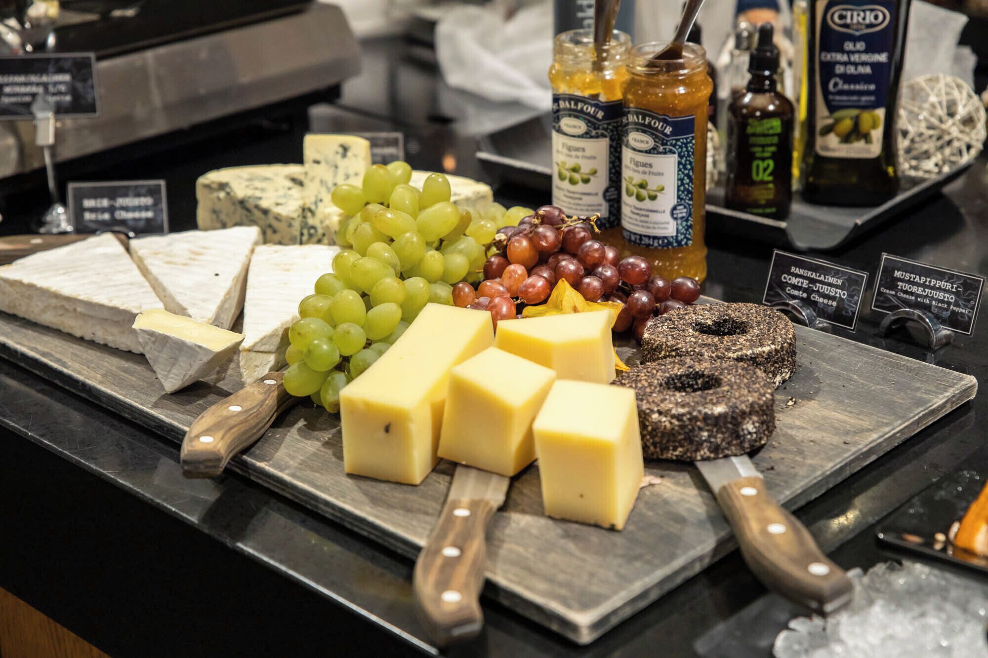 Petisqueira de queijo e frutas com diversos tipos de queijo, uvas e potes de mel sobre um balcão.