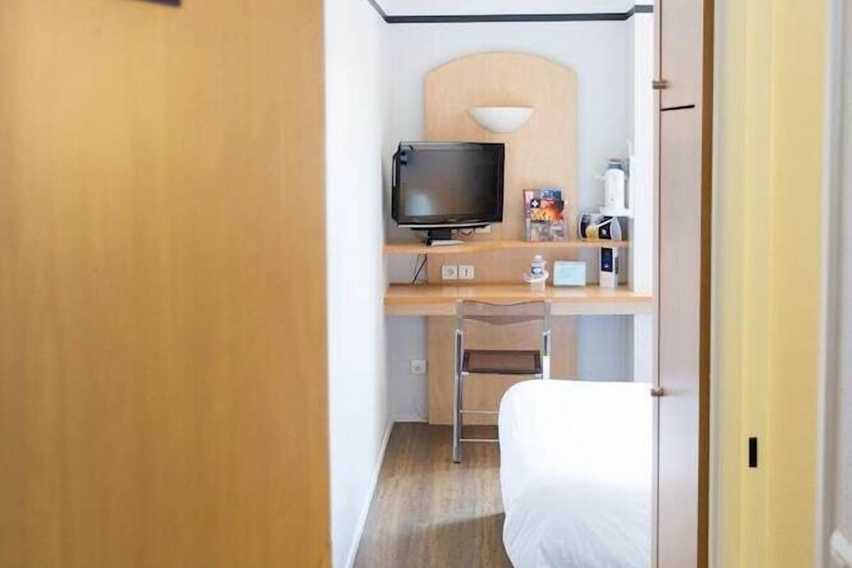 Quarto de hotel com uma cama, uma escrivaninha com televisão e uma mesinha de cabeceira com abajur.