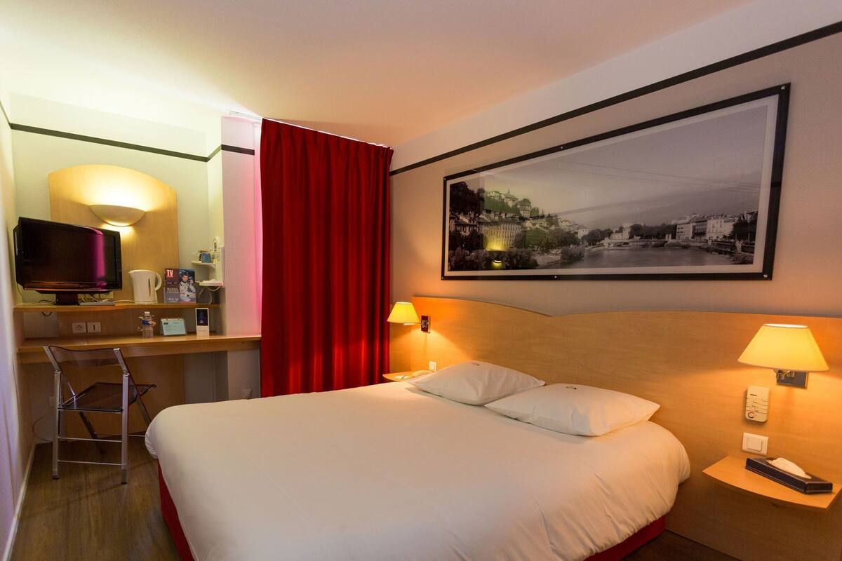 Quarto de hotel com cama, escrivaninha com computador, cadeira, televisão e um quadro na parede.