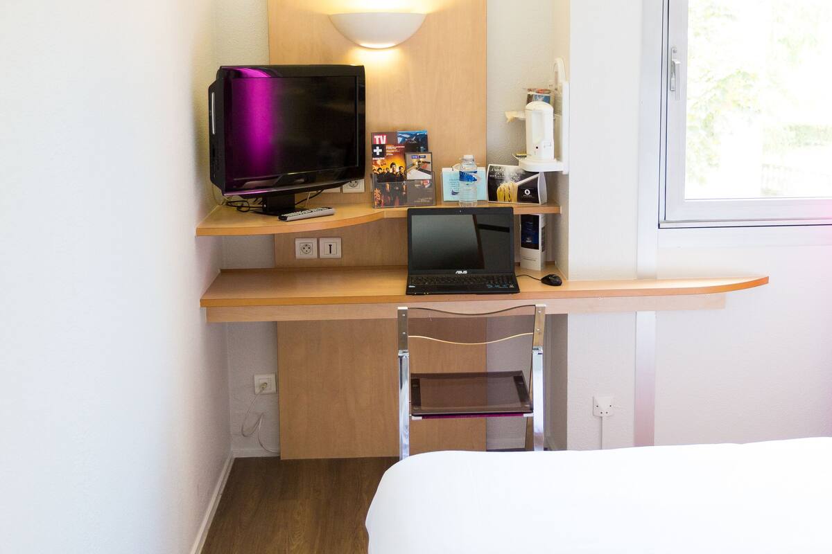 Quarto de hotel com uma cama, uma escrivaninha com um laptop, uma televisão e uma janela.