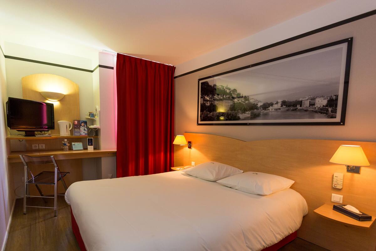 Quarto de hotel com cama, escrivaninha com computador, cadeira, televisão e pintura na parede.
