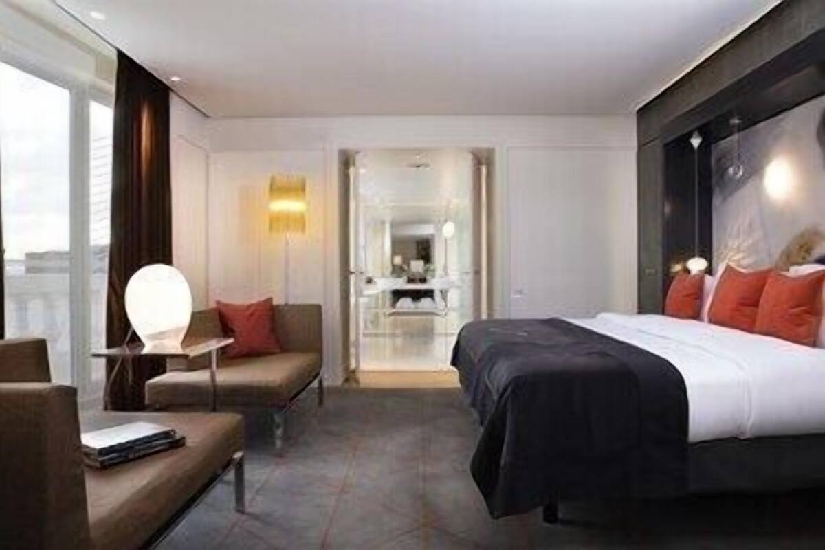 Quarto de hotel com cama, sofá, escrivaninha, espelho e banheiro visível por uma porta aberta.