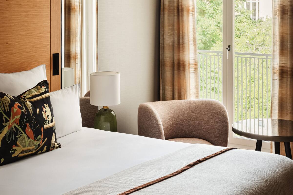 Quarto de hotel com cama, cadeira, mesa, abajur e vista para o verde lá fora.