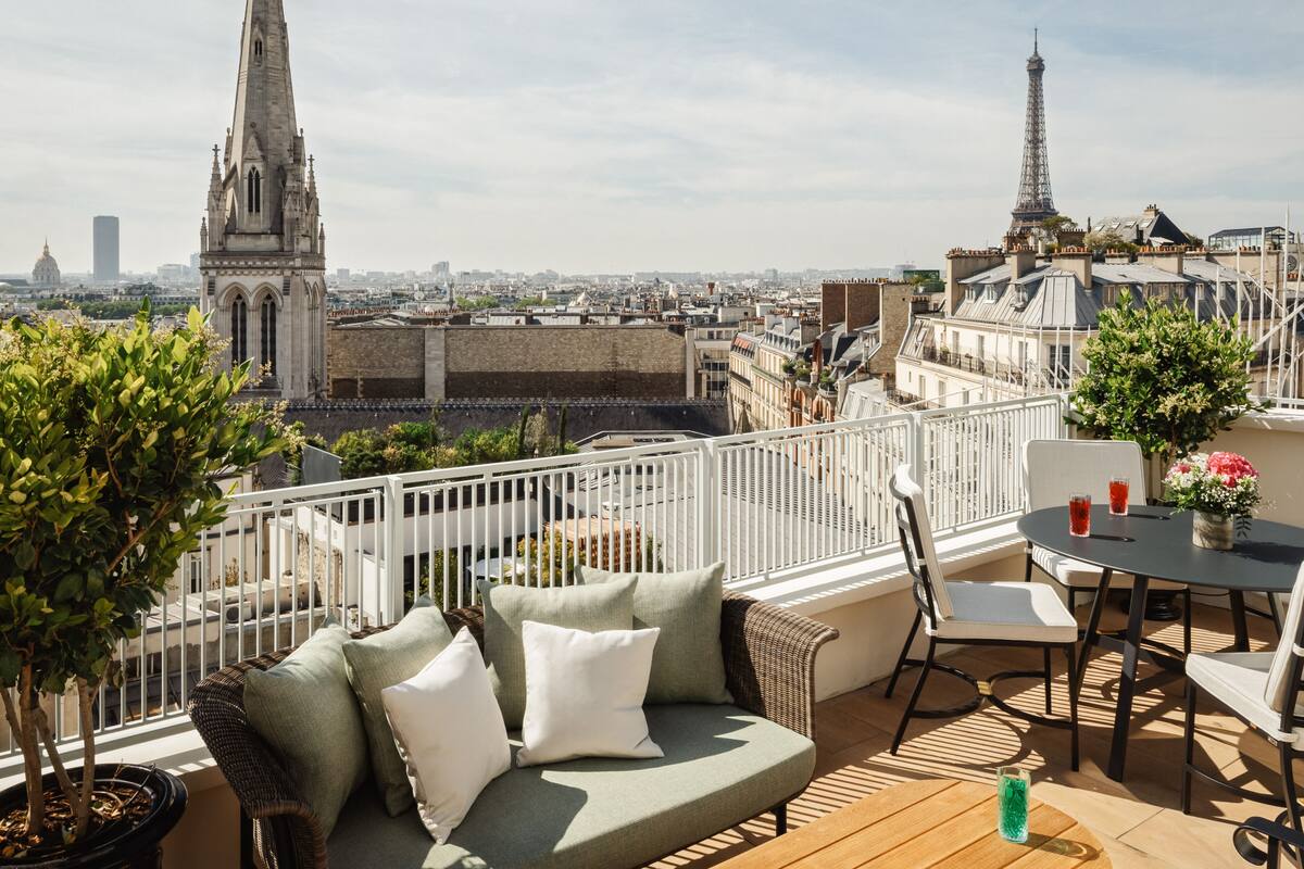 Terraço no telhado com um sofá, cadeiras e uma mesa, com vista para uma paisagem urbana com edifícios de destaque.