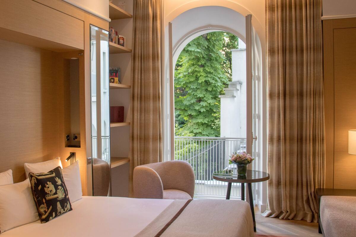Quarto de hotel com uma cama grande, uma mesa pequena com um vaso, uma cadeira e varanda com vista para vegetação.