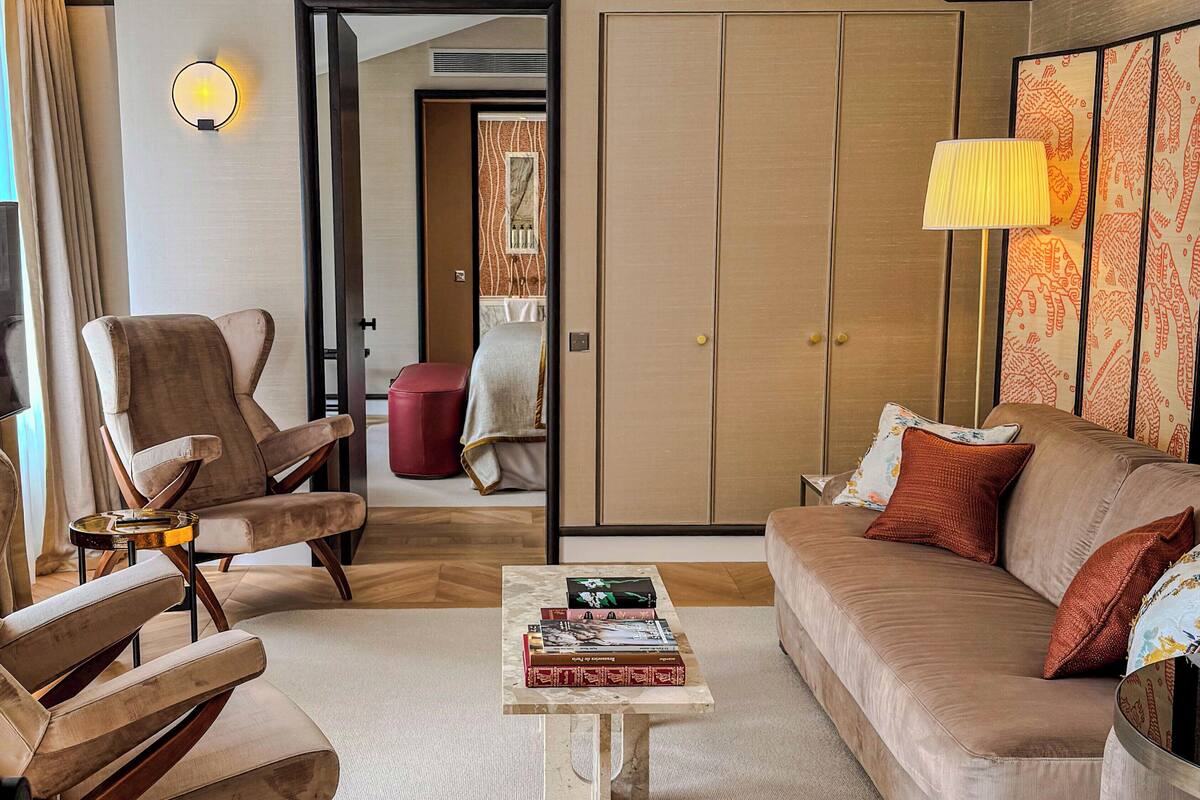 Uma sala de estar moderna com um sofá, poltrona e uma mesa de centro com livros. Há um banquinho vermelho, uma luminária e uma parede com obras de arte estampadas.
