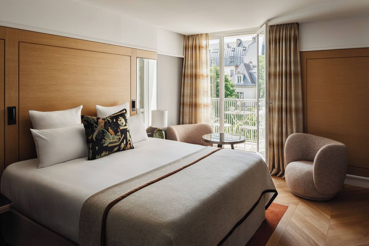 Quarto de hotel com cama grande, uma cadeira, uma mesinha e varanda com cortinas.