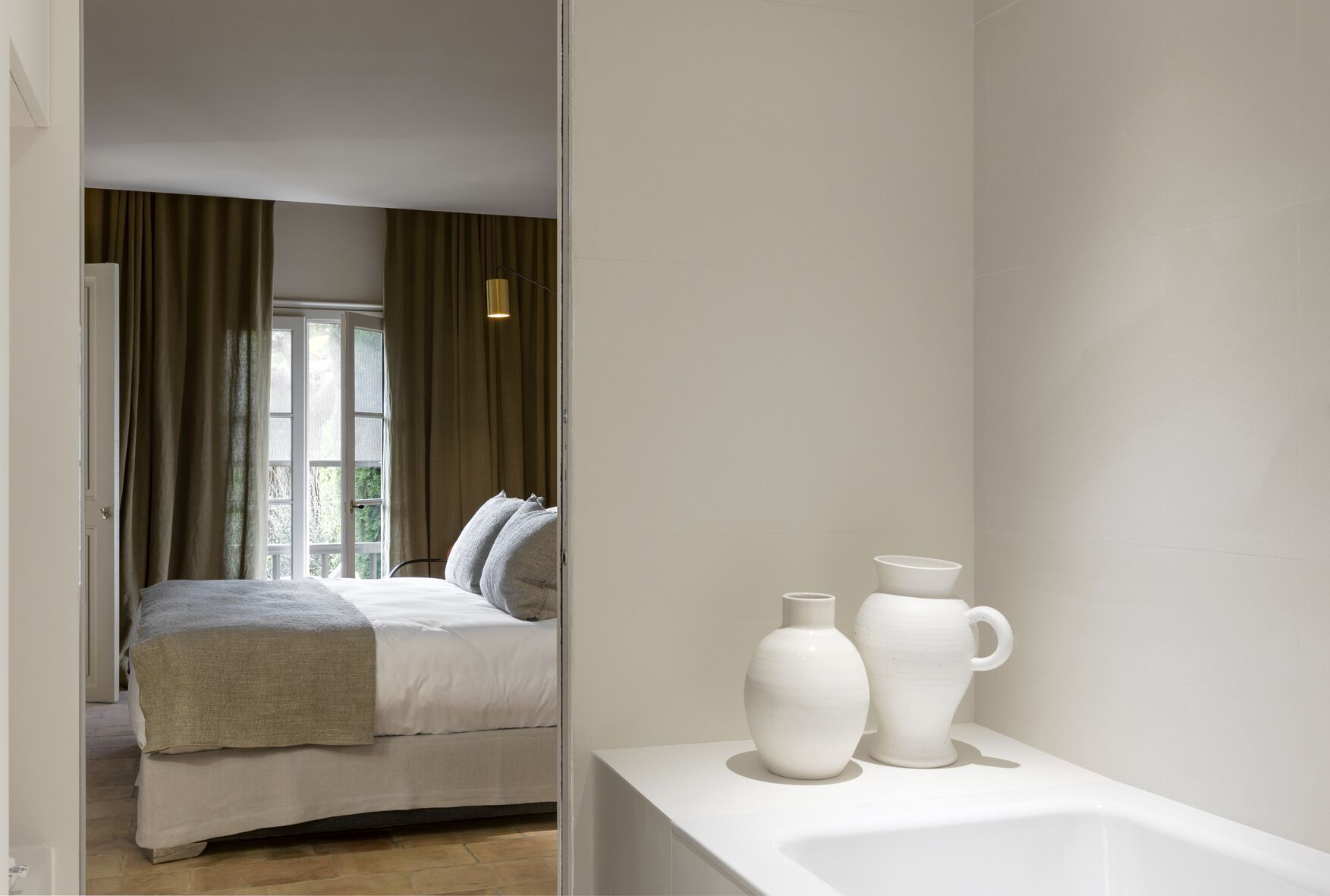 Quarto com cama, cortinas e janela. Há dois vasos brancos sobre uma superfície branca.