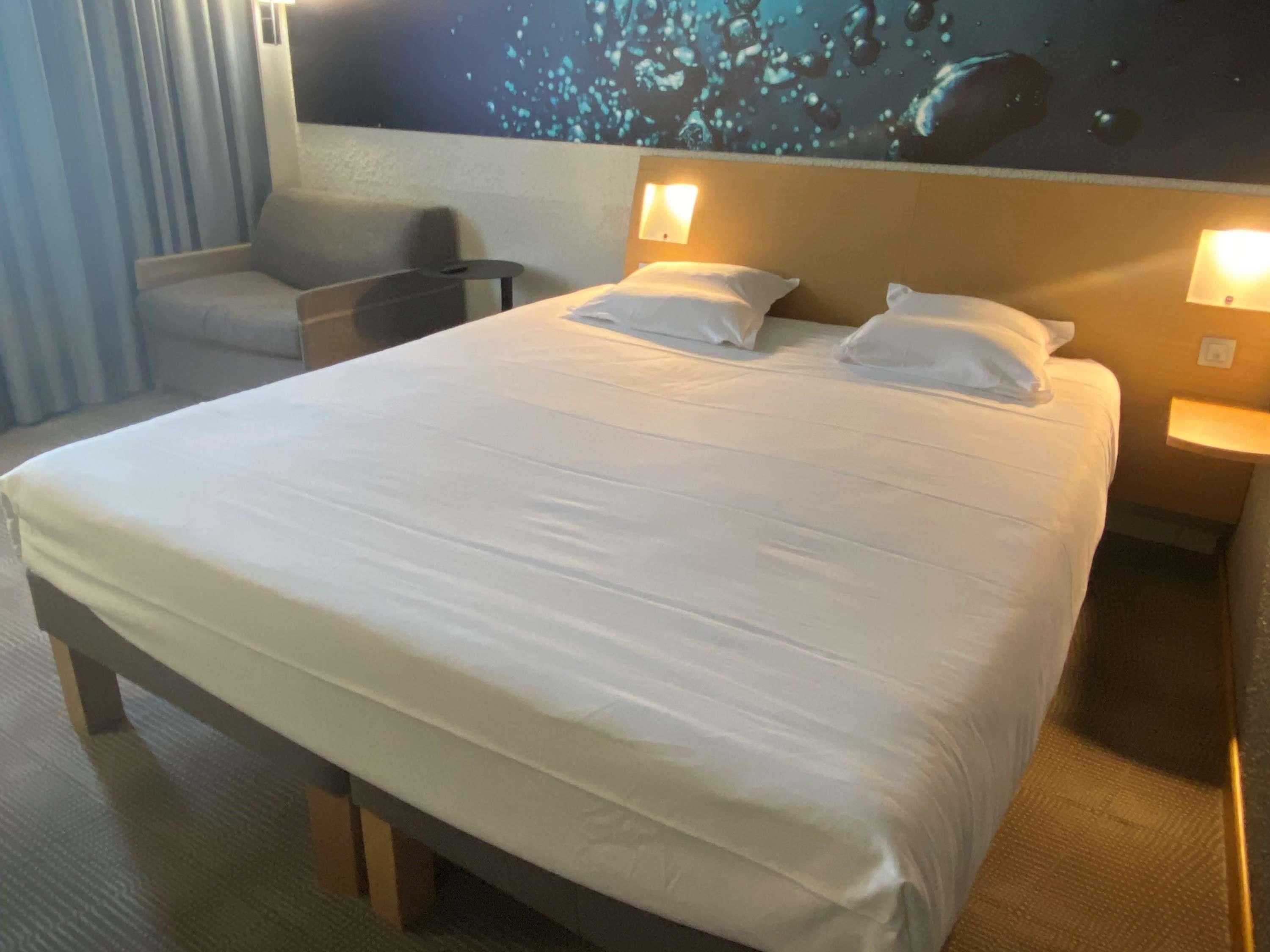 Quarto de hotel com uma cama grande, duas mesas de cabeceira com luminárias, uma cadeira e um mural na parede.