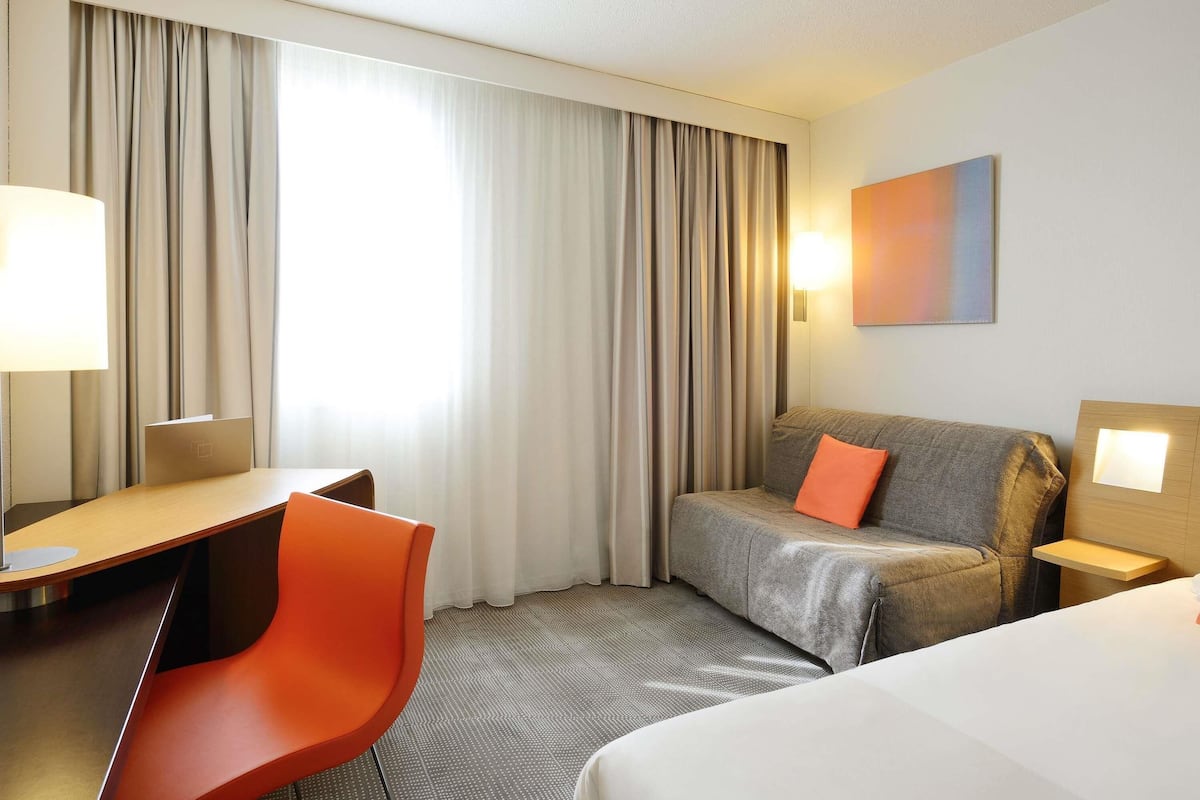 Quarto de hotel com cama, secretária com cadeira, sofá com almofada e janela com cortinas.
