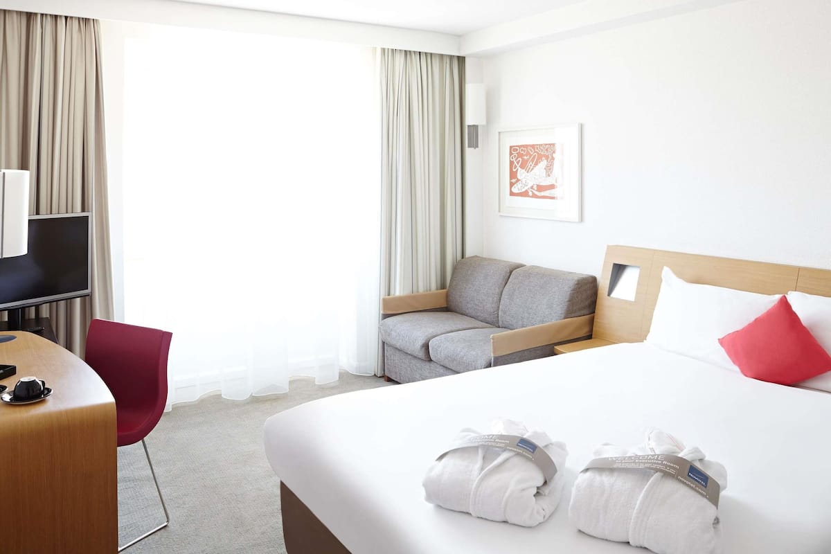 Quarto de hotel com cama, escrivaninha, cadeira, televisão e cortinas.