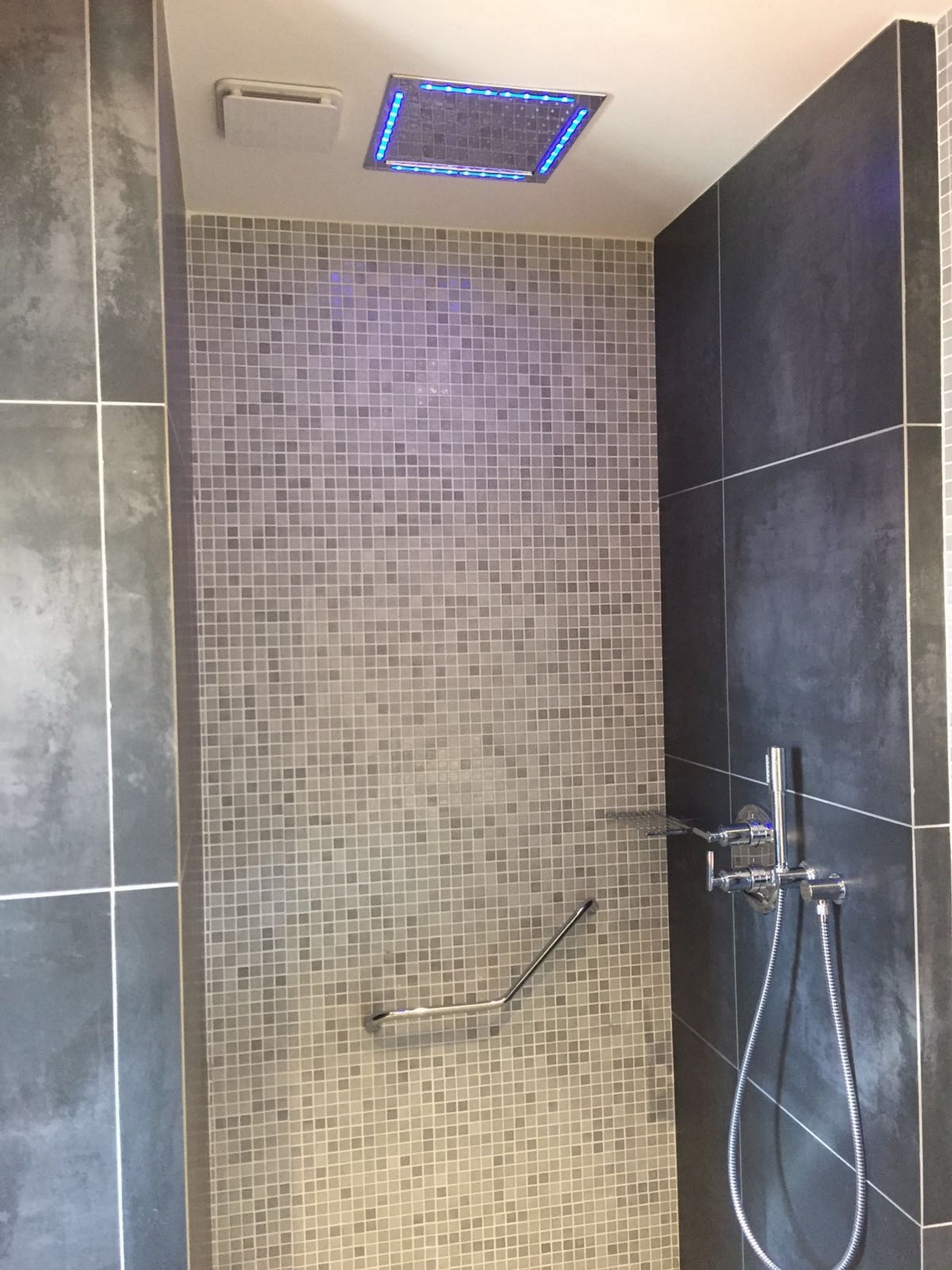 Uma área de chuveiro moderna com parede revestida de azulejos, chuveiro manual e torneira fixada na parede.