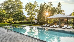 Piscina com espreguiçadeiras e um gazebo, cercada por árvores e um gramado bem cuidado.