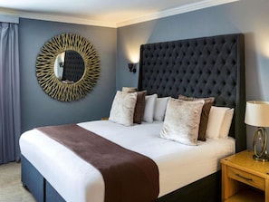 Quarto de hotel com uma cama grande, um espelho redondo, cabeceira escura e mesa de cabeceira com abajur.
