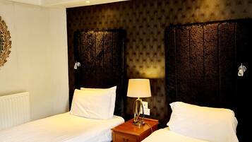 Quarto de hotel com duas camas de solteiro, uma mesa de cabeceira com abajur e cabeceira estampada escura.