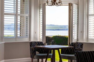 Uma área de refeições com mesa e cadeiras, janelas amplas com persianas e vista para o mar e colinas.