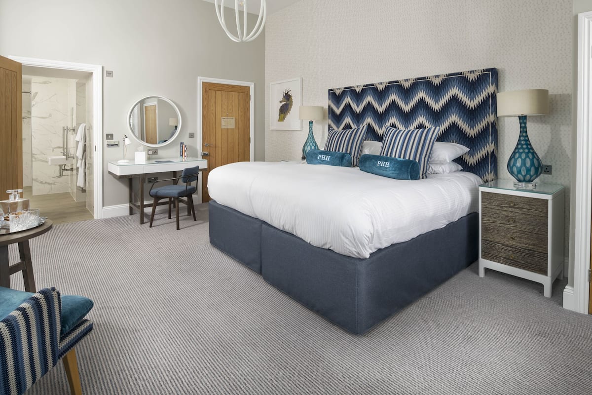 Quarto de hotel moderno com uma cama grande, uma mesa de trabalho com cadeira, um banheiro e uma mesa de cabeceira com abajur.