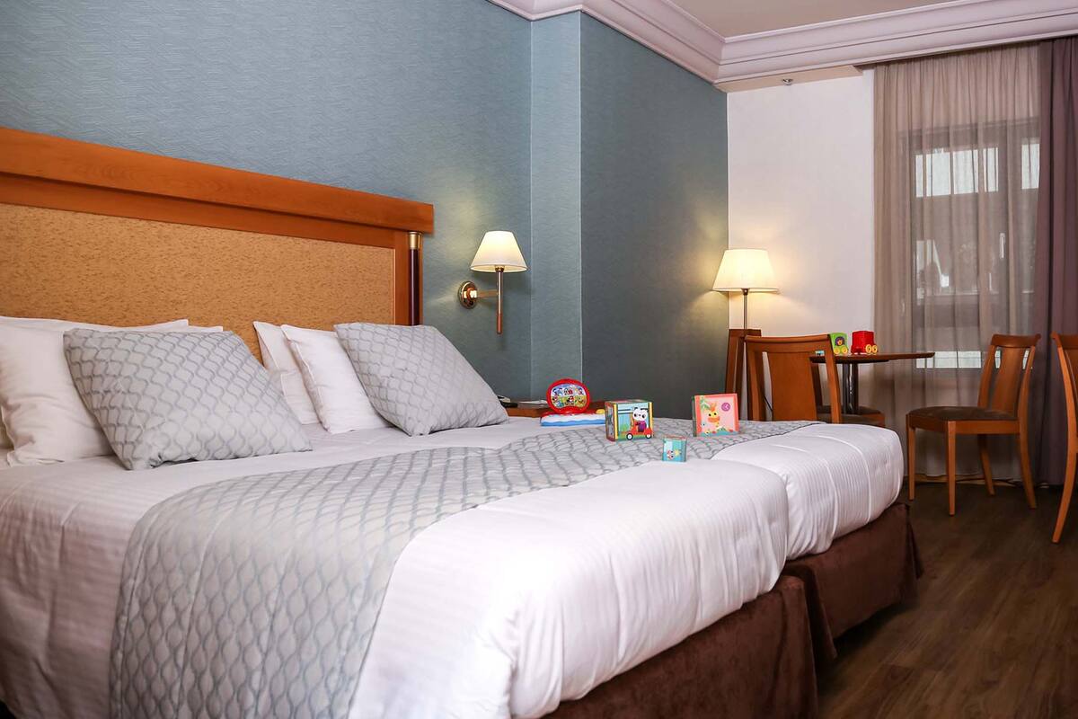 Quarto de hotel com uma cama grande, duas mesas de cabeceira, uma escrivaninha com cadeira, uma luminária e uma janela com cortinas.