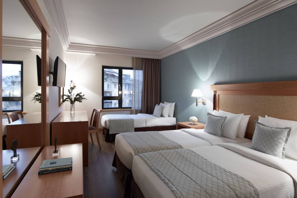 Quarto de hotel com duas camas, uma escrivaninha com cadeira, televisão, janela com vista para edifícios e abajur.
