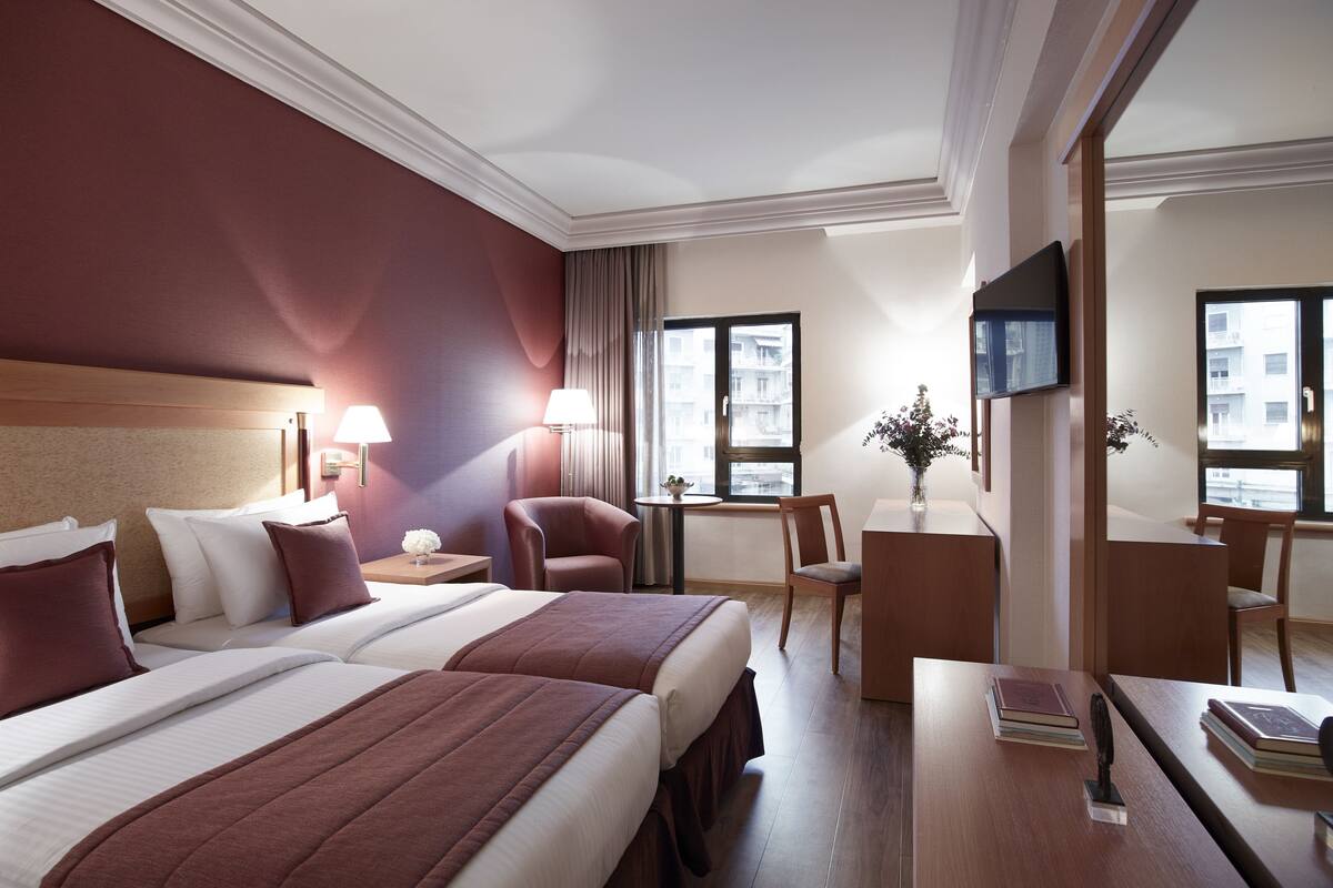 Quarto de hotel com duas camas, uma mesa, uma cadeira, uma televisão e uma janela com vista para edifícios.