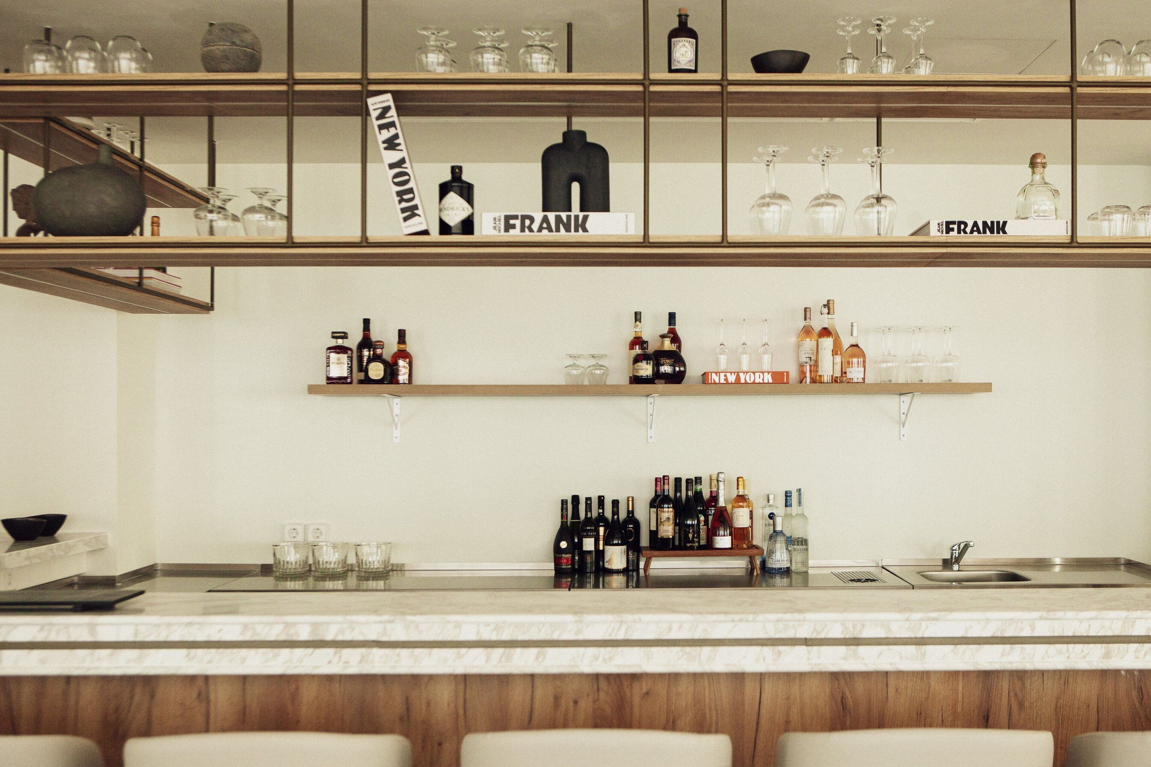 Uma área de bar com prateleiras cheias de garrafas de bebidas e copos, bancada de mármore e balcão de madeira.